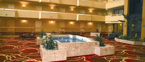 Um saguão de hotel com uma grande piscina interna, várias áreas de estar e um teto alto com iluminação embutida.