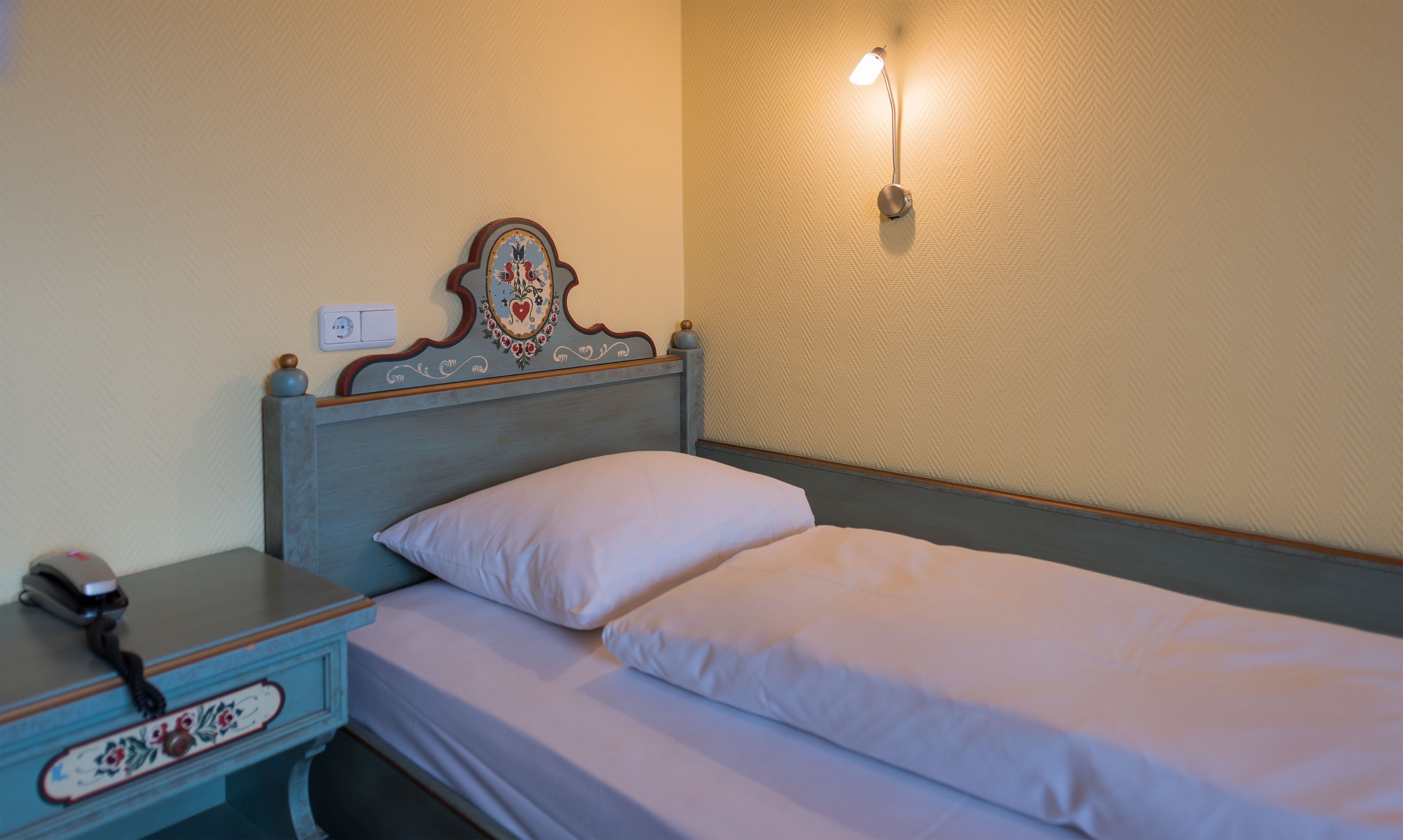 Uma cama de solteiro com roupa de cama branca, cabeceira de madeira com uma decoração ornamental, mesa de cabeceira com telefone e luminária fixa na parede.