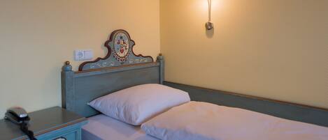 Uma cama de solteiro com roupa de cama branca, cabeceira de madeira com uma decoração ornamental, mesa de cabeceira com telefone e luminária fixa na parede.