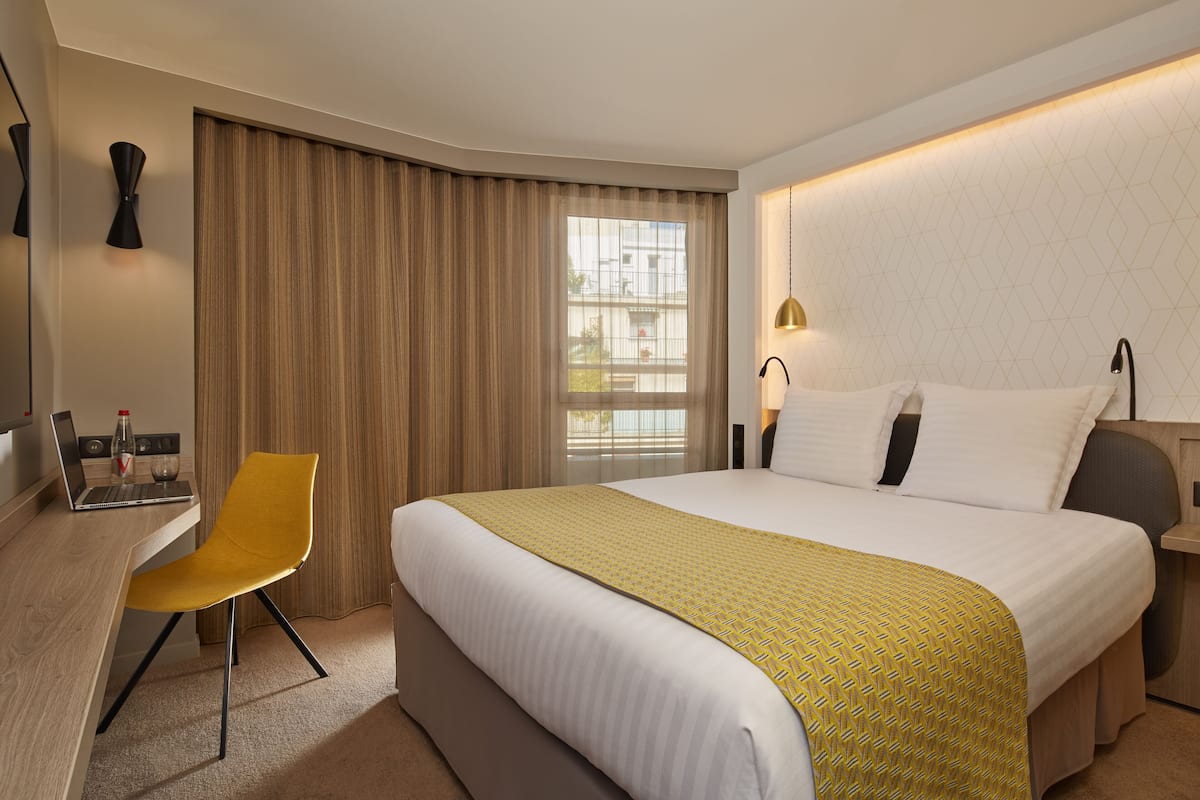 Quarto de hotel com uma cama grande, uma escrivaninha com um laptop, uma cadeira amarela e uma janela com cortinas.