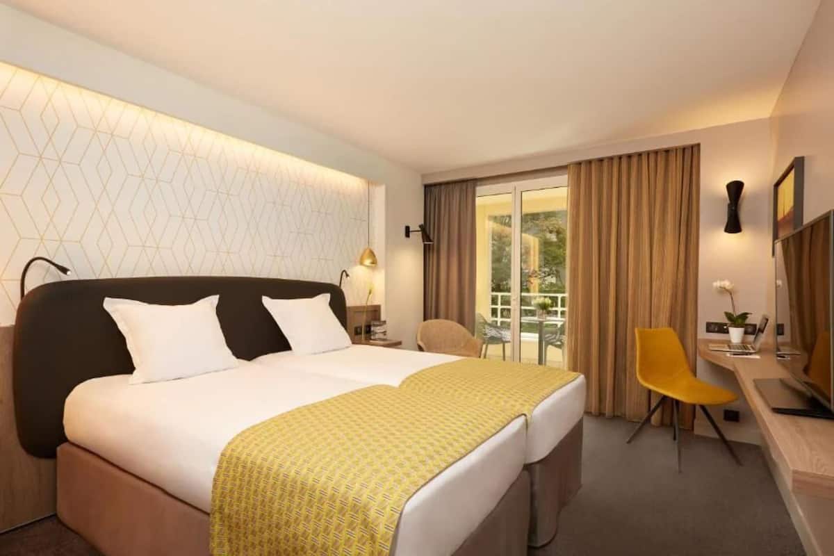 Quarto de hotel com uma cama grande, uma escrivaninha com cadeira, um laptop e vista para o exterior através de uma porta de correr.