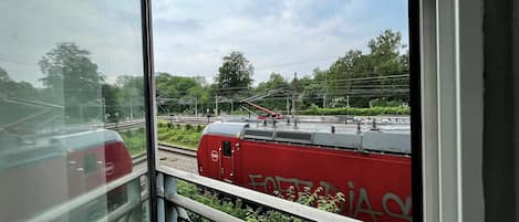 Plataforma de estação de trem com um trem vermelho nos trilhos, céu claro e vegetação ao fundo.