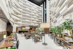 Um átrio moderno de hotel com várias áreas de refeições, tetos altos e plantas em vasos.