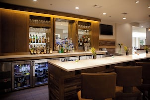Uma área de bar com um balcão comprido, várias prateleiras com garrafas, copos e uma televisão na parede.