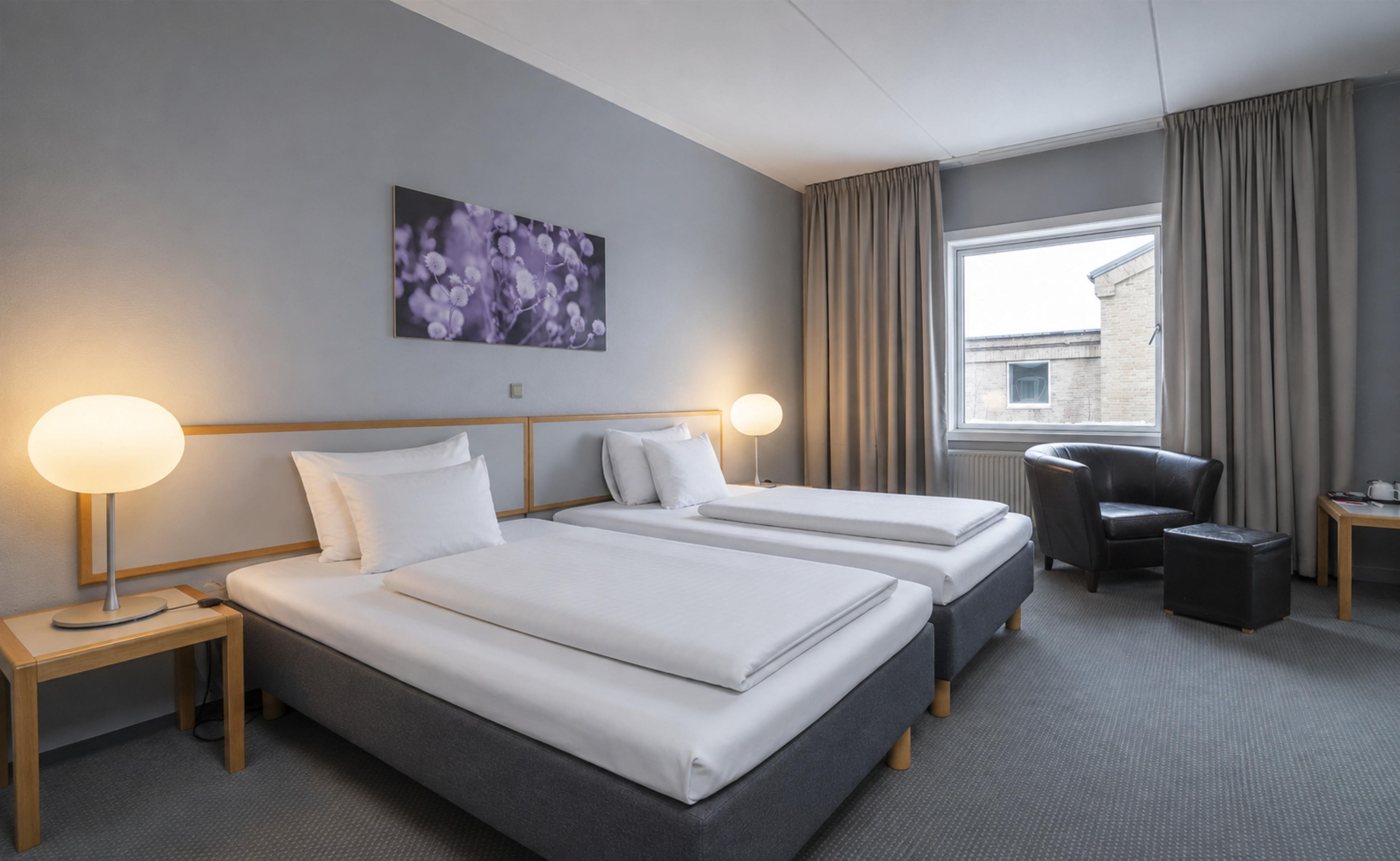 Quarto de hotel com duas camas, uma mesa, uma cadeira e uma janela com cortinas.