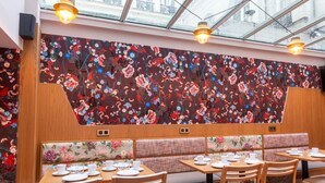 Um restaurante com bancos e mesas de madeira, um mural floral na parede e um claraboia acima.