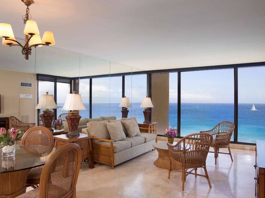 Uma sala de estar espaçosa com uma grande janela com vista para o oceano, mobiliada com cadeiras de vime, um sofá e uma mesa de centro de vidro.