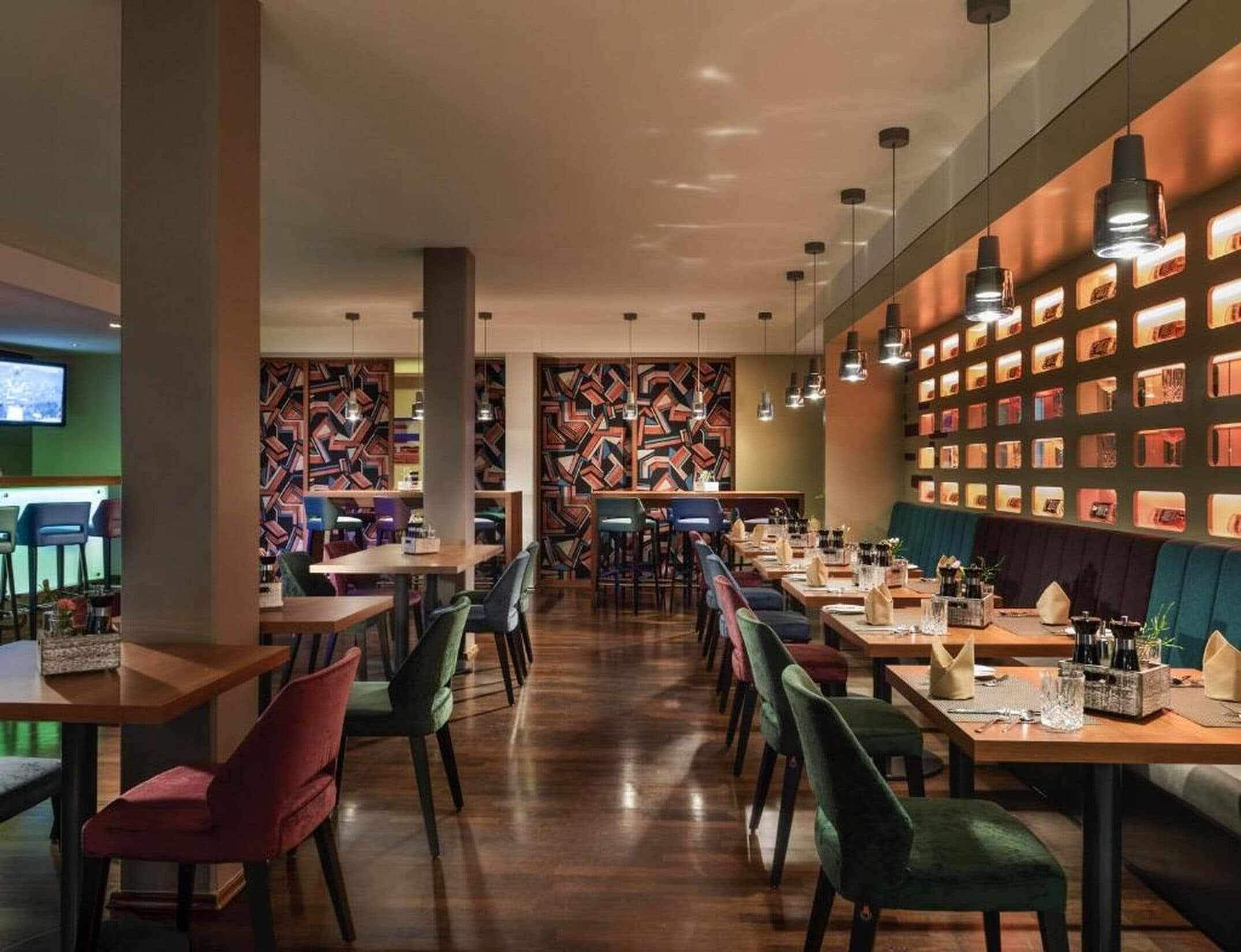 Um restaurante com um balcão comprido, várias mesas e cadeiras. Há luminárias pendentes e uma parede com padrão ao fundo.