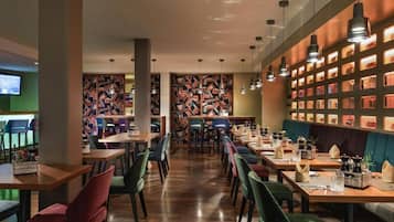 Um restaurante com um balcão comprido, várias mesas e cadeiras. Há luminárias pendentes e uma parede com padrão ao fundo.