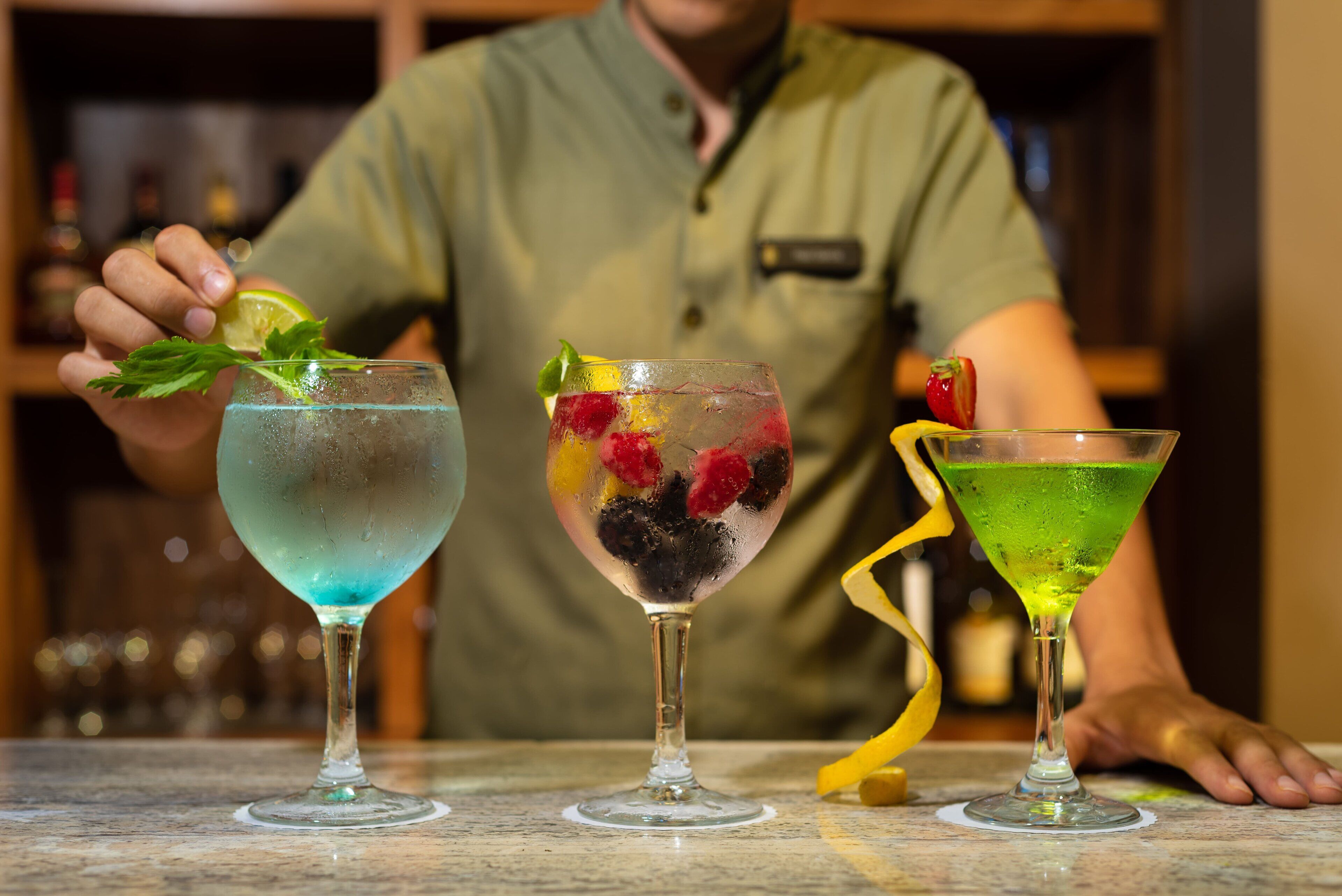 Três cocktails sobre um balcão de bar, com um bartender ao fundo.