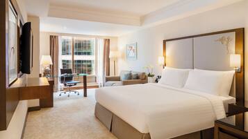 Quarto de hotel com uma cama grande, uma escrivaninha com cadeira, um sofá e uma televisão na parede.