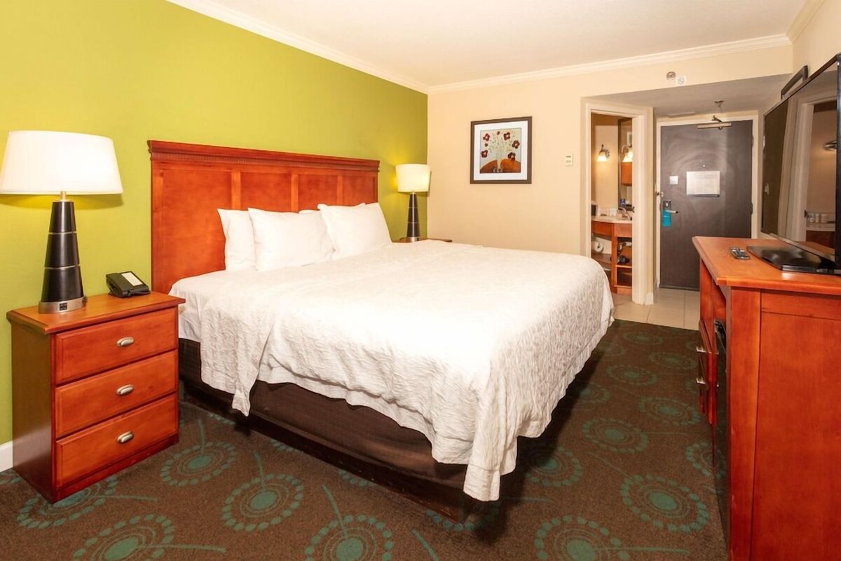 Quarto de hotel com uma cama grande, mesa de cabeceira de madeira, escrivaninha e espelho.