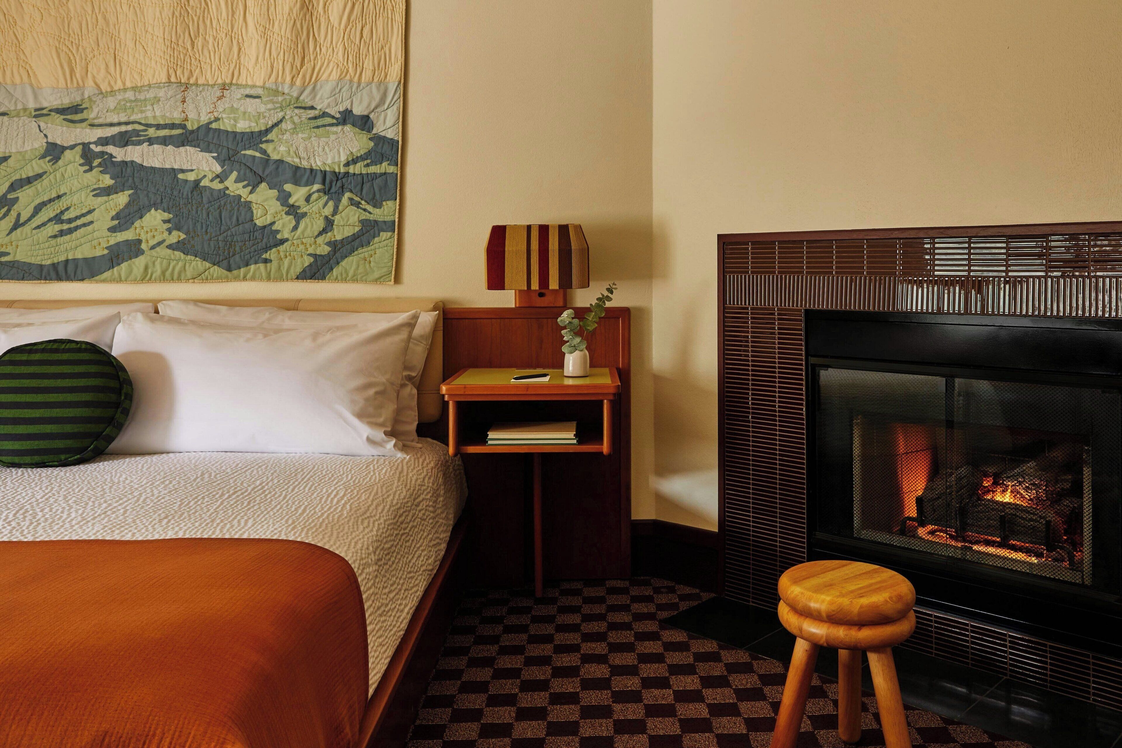 Quarto de hotel com uma cama, uma mesinha de cabeceira, uma pequena mesa com um vaso e um livro, chão quadriculado e lareira.