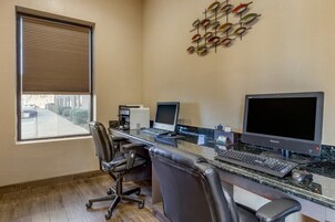 A hotel office with a desktop computer, a laptop, and a monitor. There is a window with blinds, a wall-mounted decorative item, and a chair.