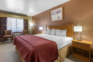 Quarto de hotel com uma cama grande, uma mesa com cadeira, mesa de cabeceira com abajur e um quadro na parede.