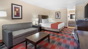 Quarto de hotel com sofá cinzento, uma cama simples com manta vermelha, mesa de centro de madeira e tapete com padrão em vermelho e preto.