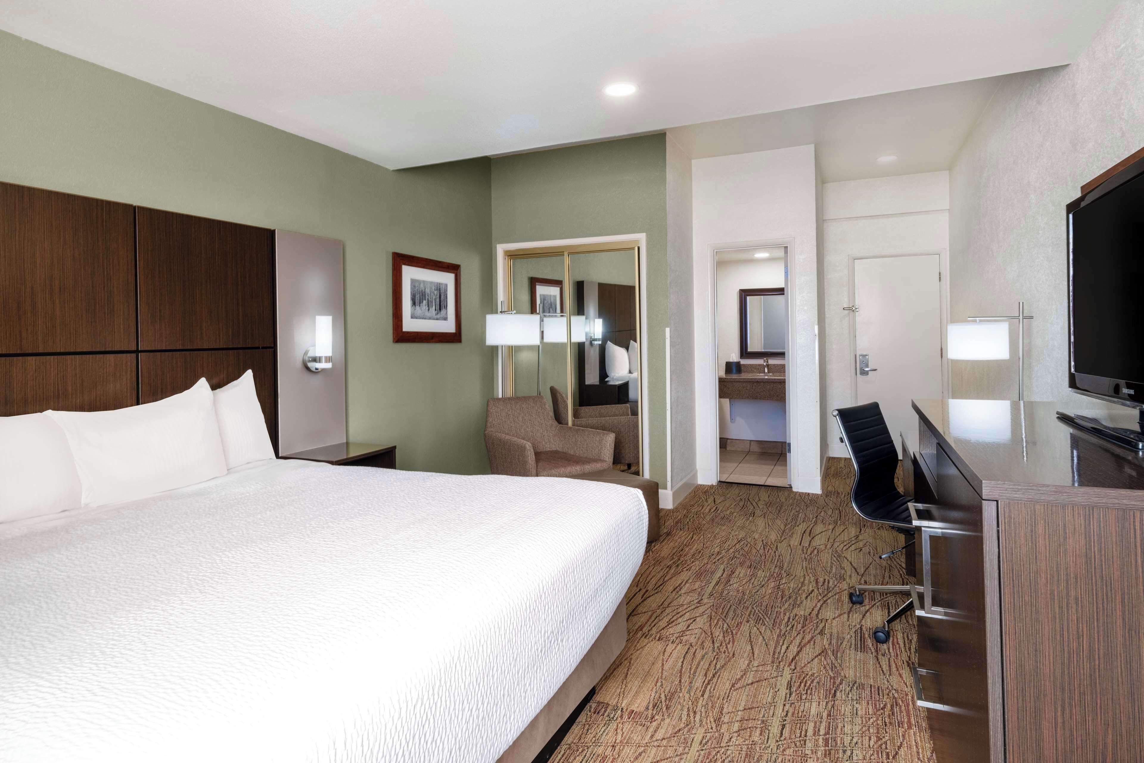 Quarto de hotel com uma cama grande, uma escrivaninha com cadeira, televisão e banheiro visível através de uma porta aberta.