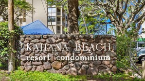Placa do resort e condomínio KAHANA BEACH, com um muro de pedra e um telhado azul visíveis ao fundo.