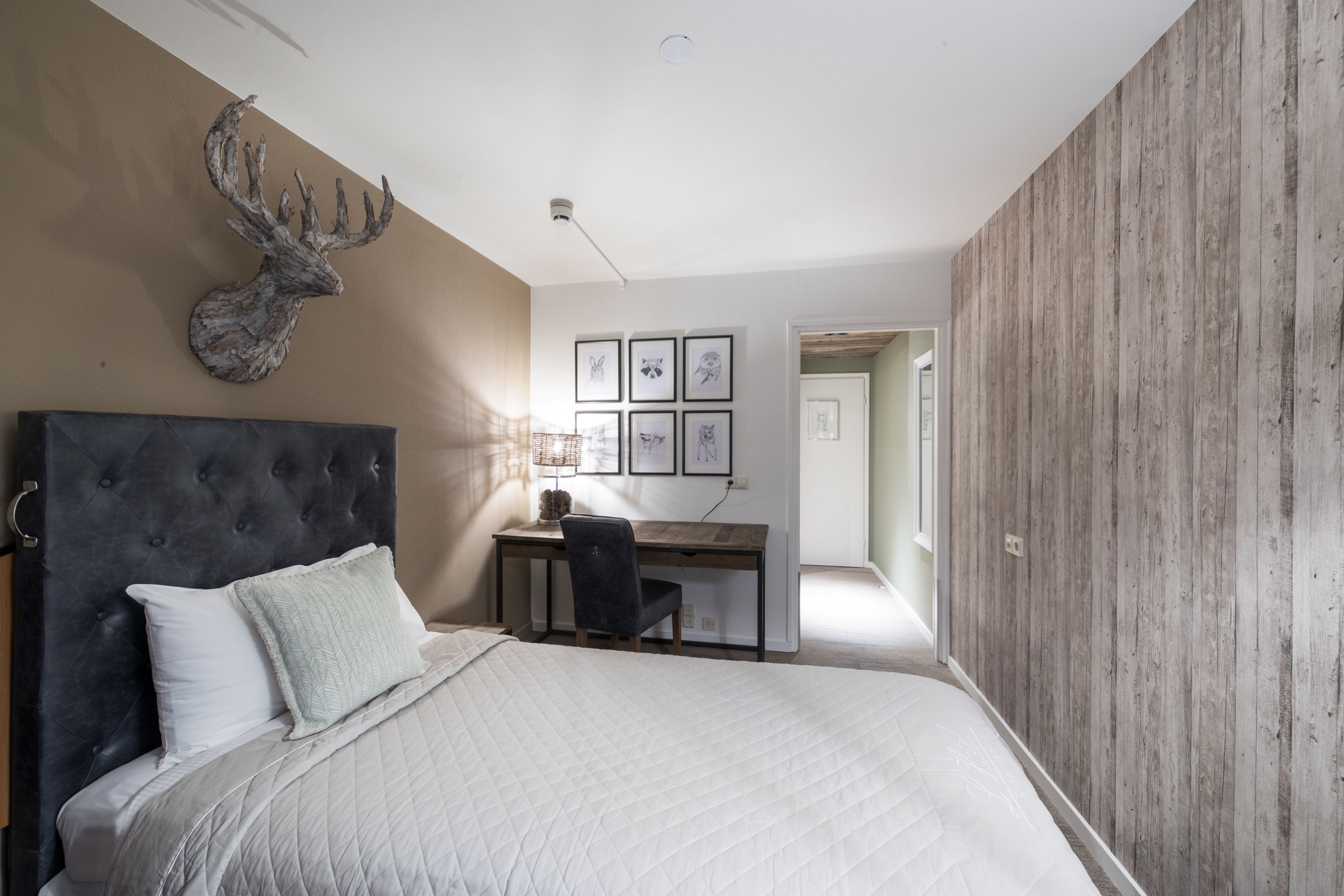 Quarto com cama grande, escrivaninha e cadeira. Há um chifre de veado como decoração na parede e quadros enquadrados.