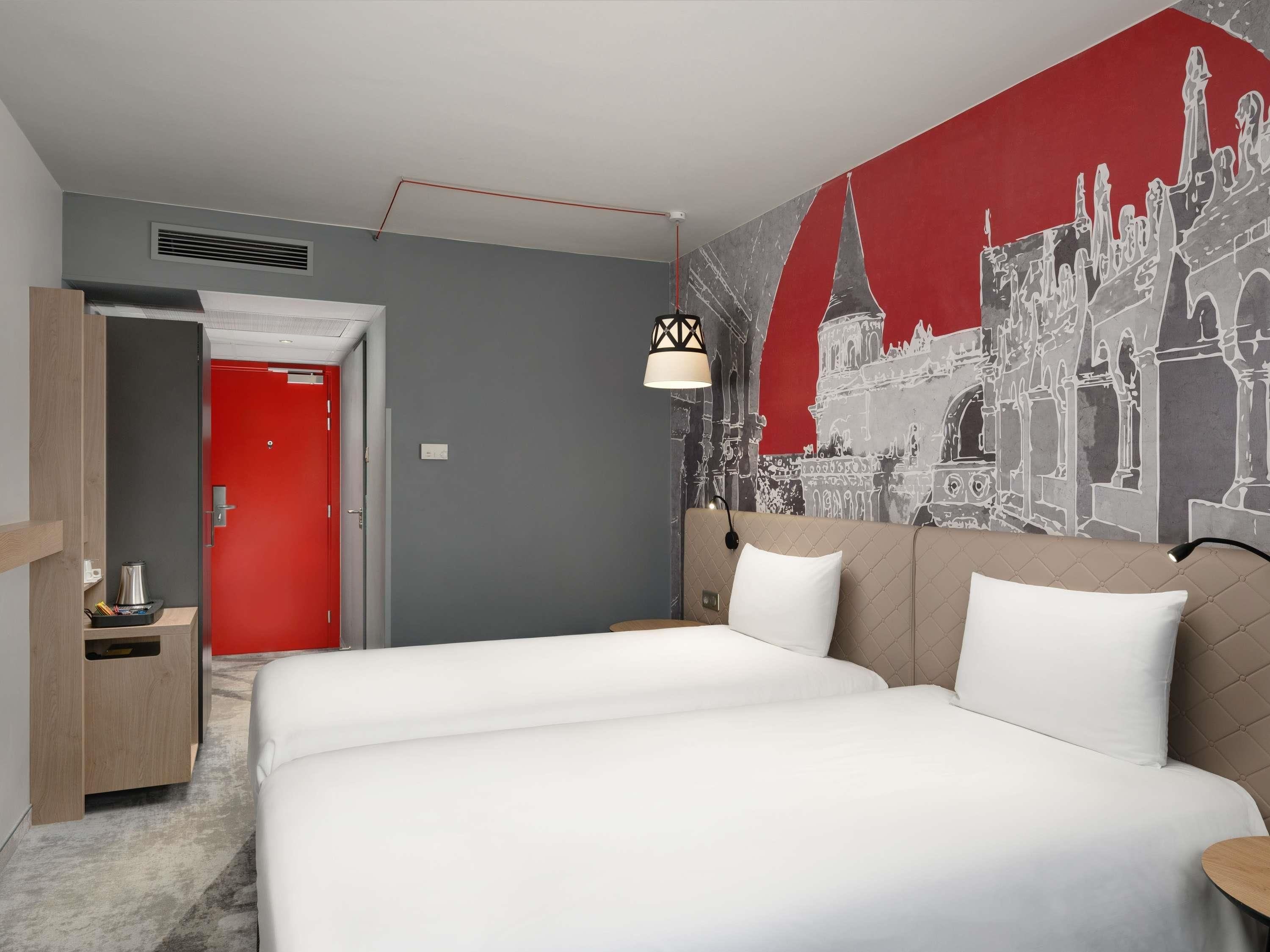 Quarto de hotel com cama, porta vermelha, painel na parede e mesa de cabeceira.