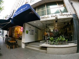 Entrada de hotel com fachada de vidro, área de estar externa e placa com a mensagem 'Boa viagem'.