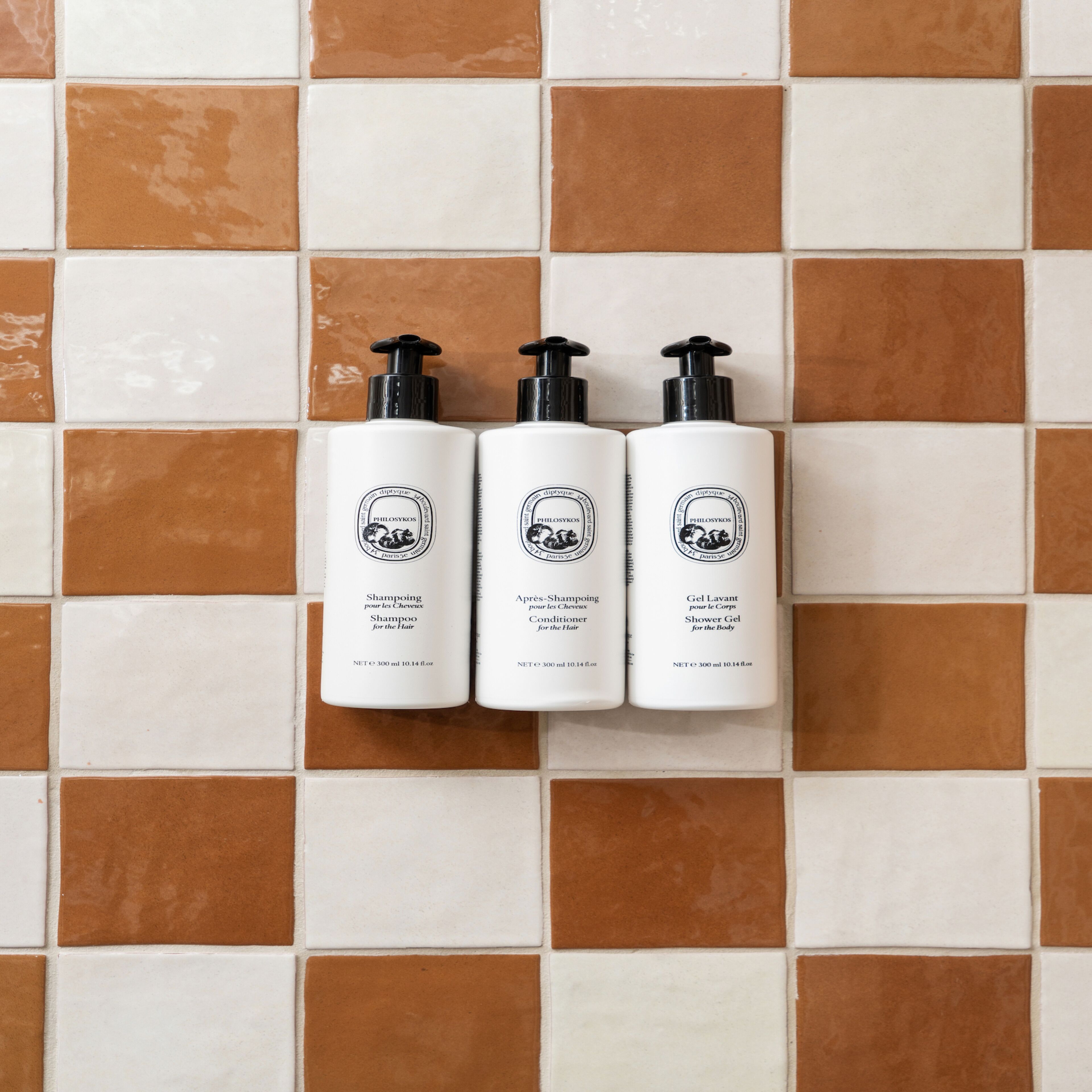 Três garrafas brancas com bombas e etiquetas pretas, dispostas numa parede com azulejos.