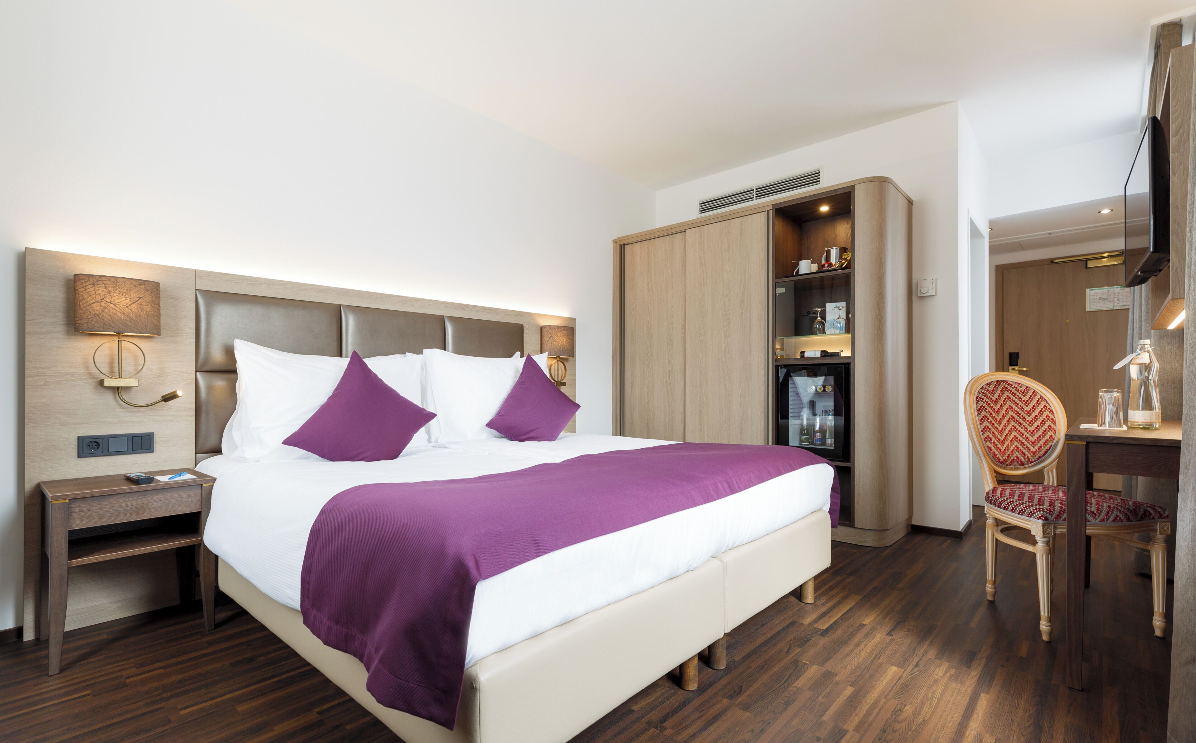 Quarto de hotel com cama, mesa de cabeceira, guarda-roupa, escrivaninha, cadeira e televisão.