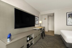 Quarto de hotel com televisão de tela plana, escrivaninha com luminária, cama e minibar.