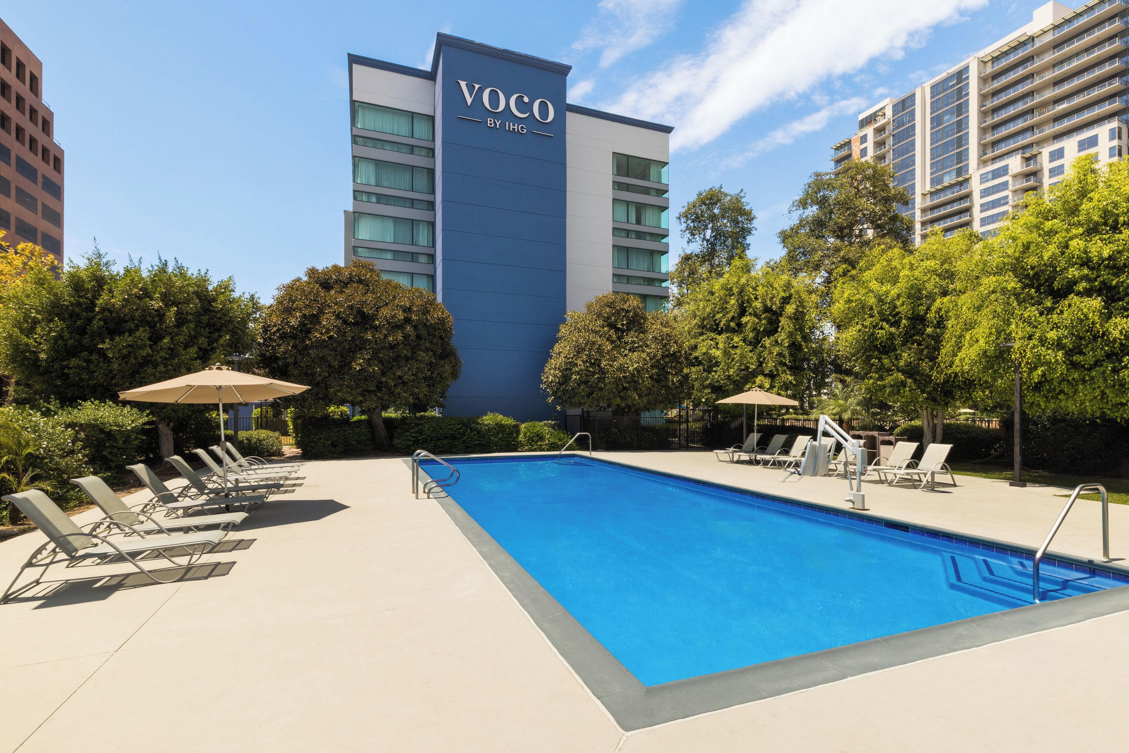 Piscina com espreguiçadeiras e guarda-sóis, rodeada por árvores e um edifício com a identificação 'Voco by IHG'.