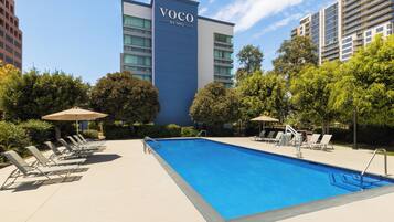 Piscina com espreguiçadeiras e guarda-sóis, rodeada por árvores e um edifício com a identificação 'Voco by IHG'.