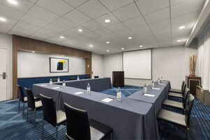 Uma sala de conferências com uma mesa comprida, cadeiras, tela de projeção e um orador.