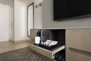 Quarto de hotel moderno com televisão de tela plana na parede, cafeteira e uma pequena mesa com uma xícara branca e cápsulas de café.
