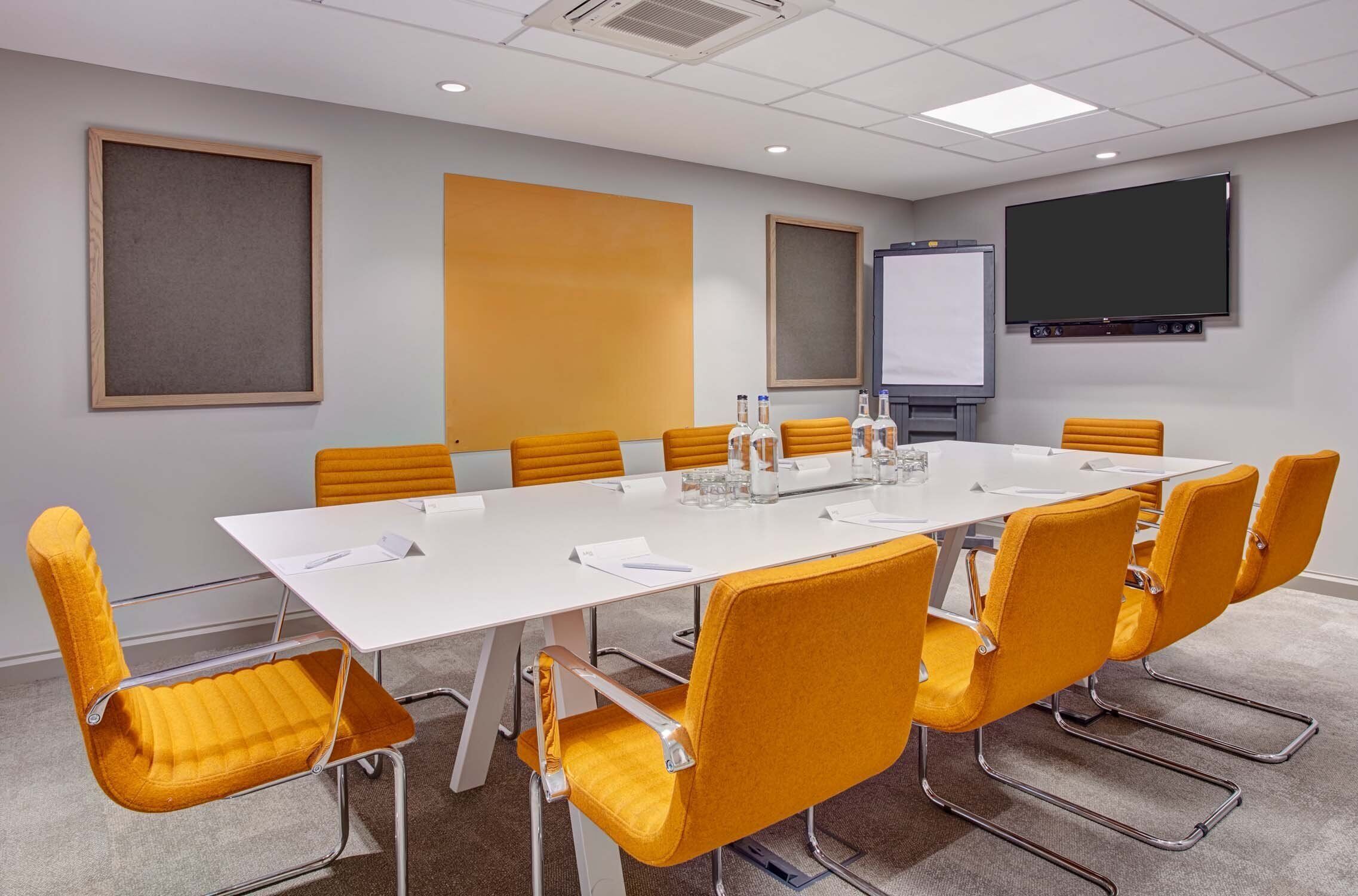 Uma sala de conferências com uma mesa branca, cadeiras laranja, uma tela de projetor e um quadro branco.