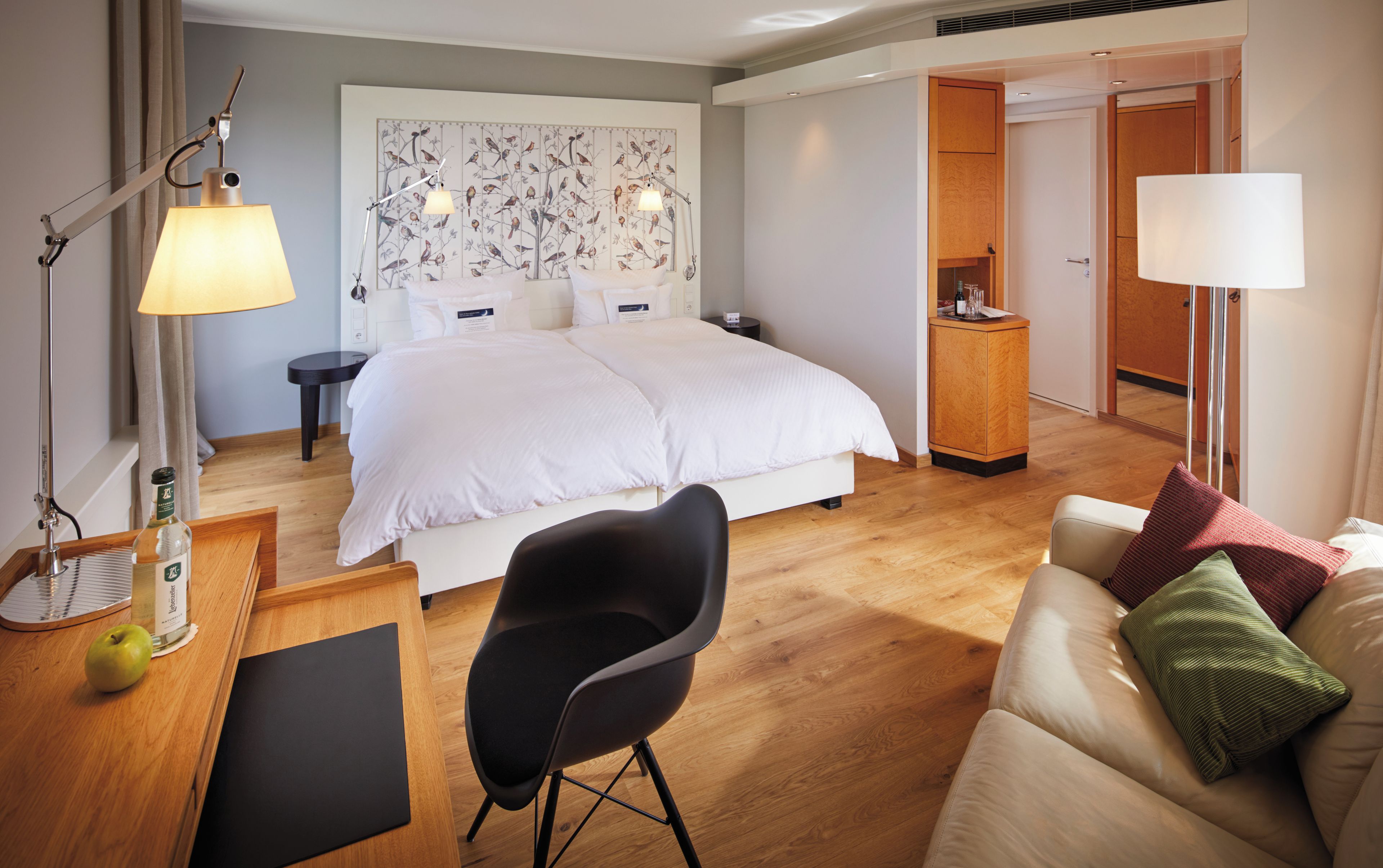 Quarto de hotel com uma cama grande, uma escrivaninha com abajur, uma cadeira, um sofá e um armário de madeira.
