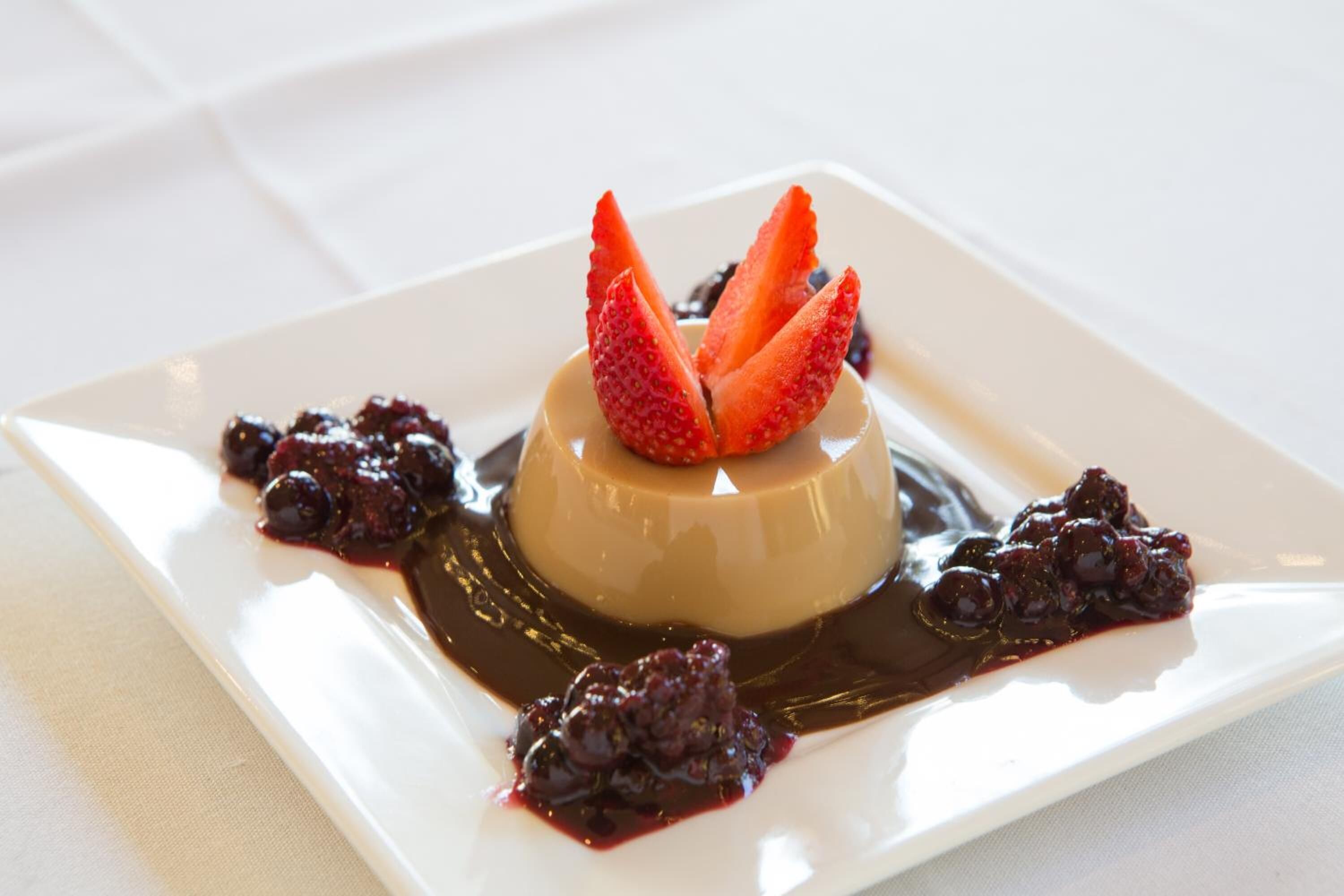 Sobremesa em um prato branco com base quadrada, coberta com uma morango e rodeada por frutas vermelhas e calda de chocolate.