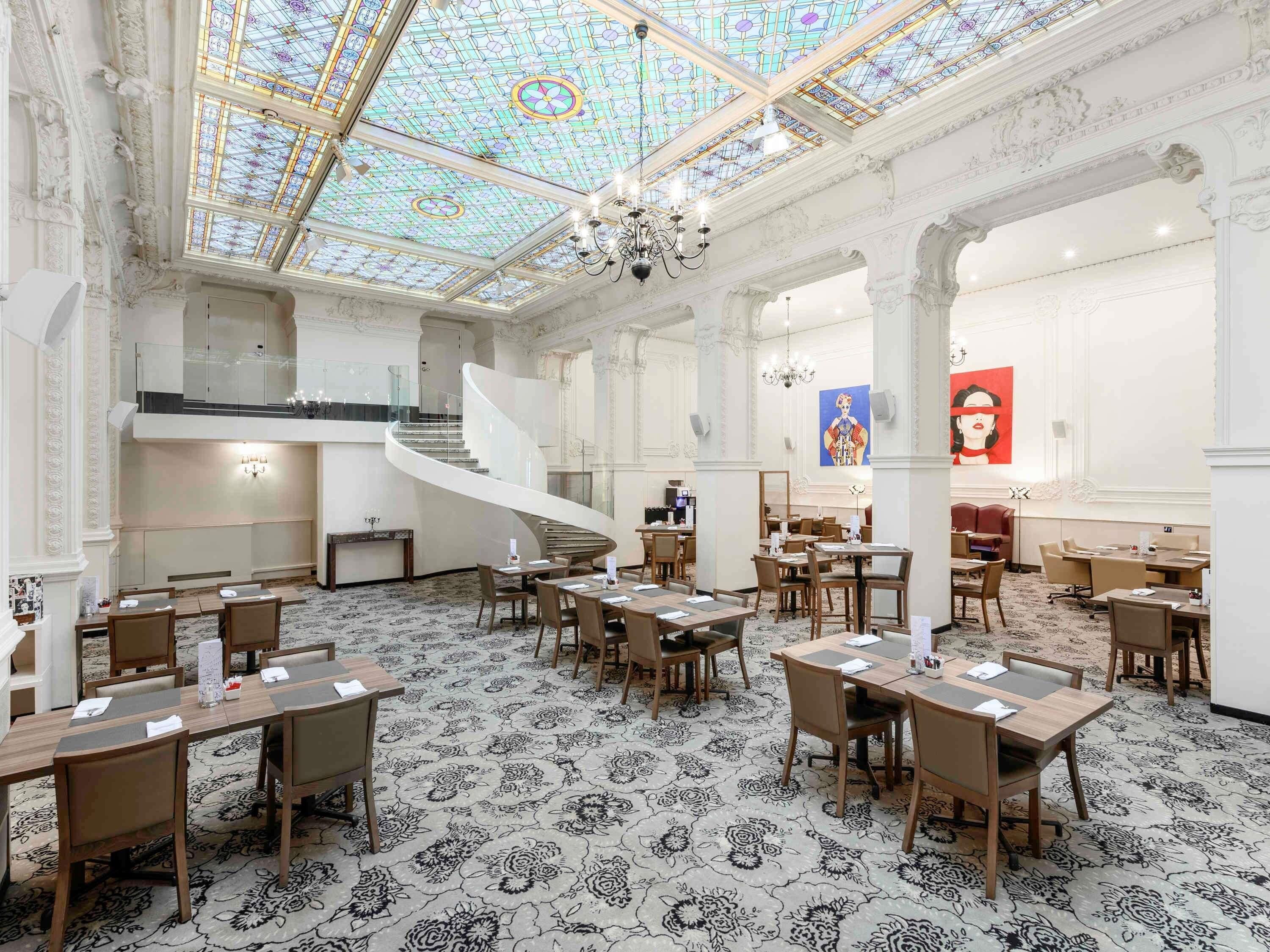 Um amplo saguão de hotel com uma escadaria central, várias áreas de refeições e um teto de vitrais.