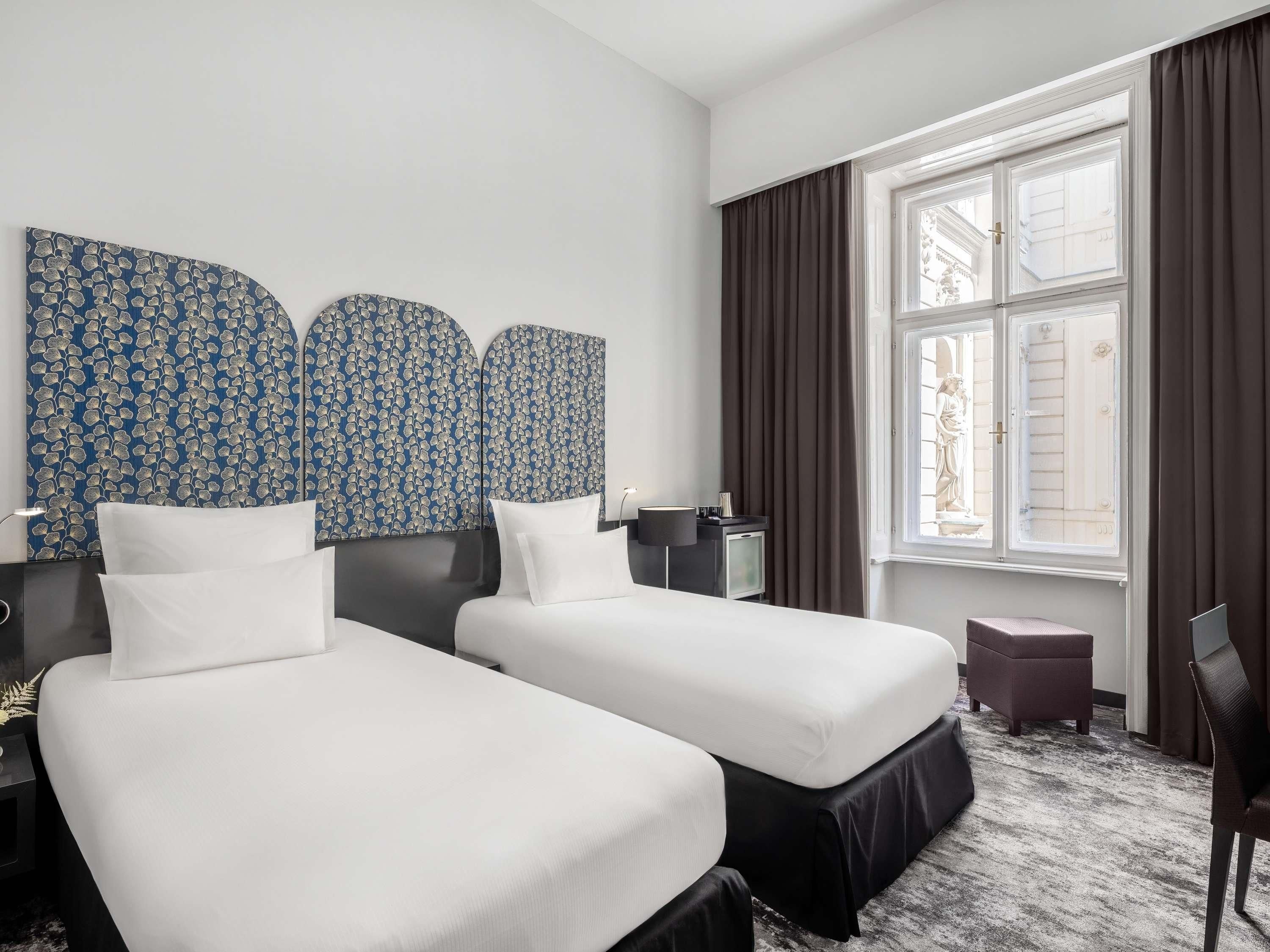 Quarto de hotel com duas camas, uma janela grande com cortinas, uma mesinha com abajur e um painel decorativo na parede.