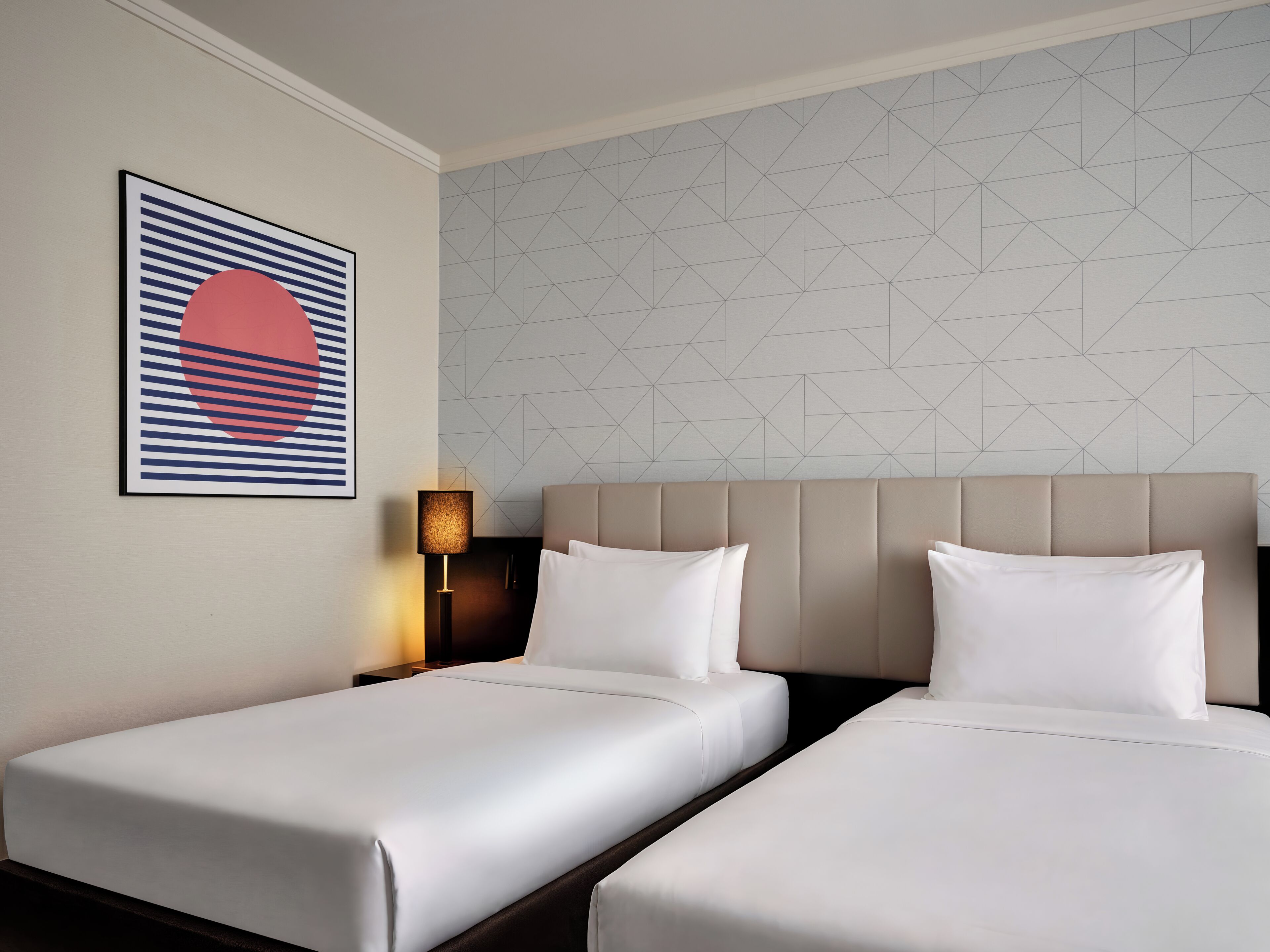 Quarto de hotel com duas camas, uma obra de arte pendurada na parede, parede com padrão geométrico e uma luminária de cabeceira.