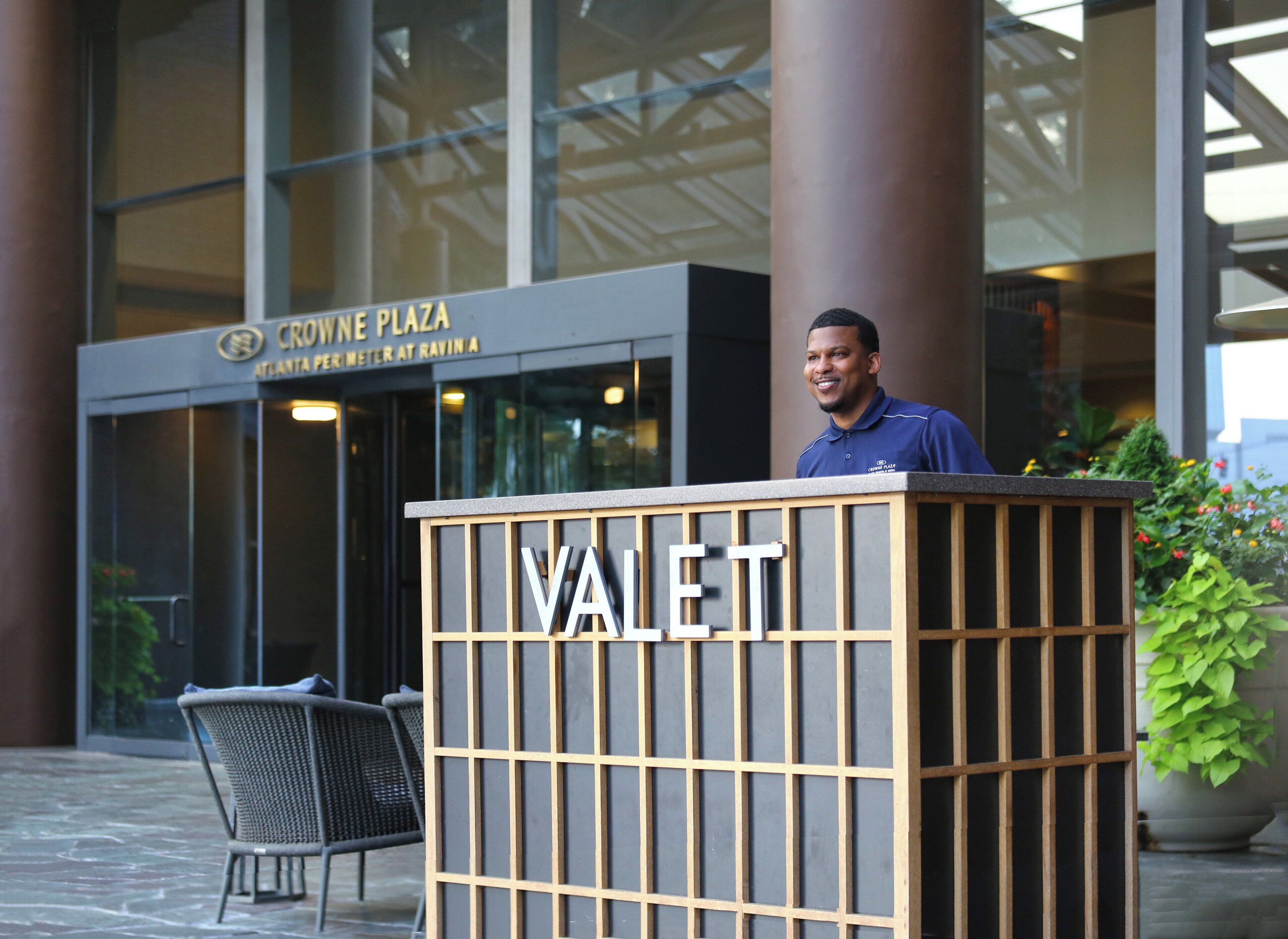 Um homem está em um pódio com a palavra "VALET" em frente a um edifício identificado como "CROWNE PLAZA".