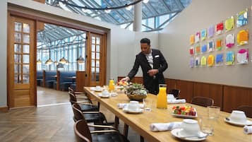 Uma sala de jantar com uma longa mesa de madeira posta para o café da manhã, uma cesta de frutas e um copo de suco de laranja.