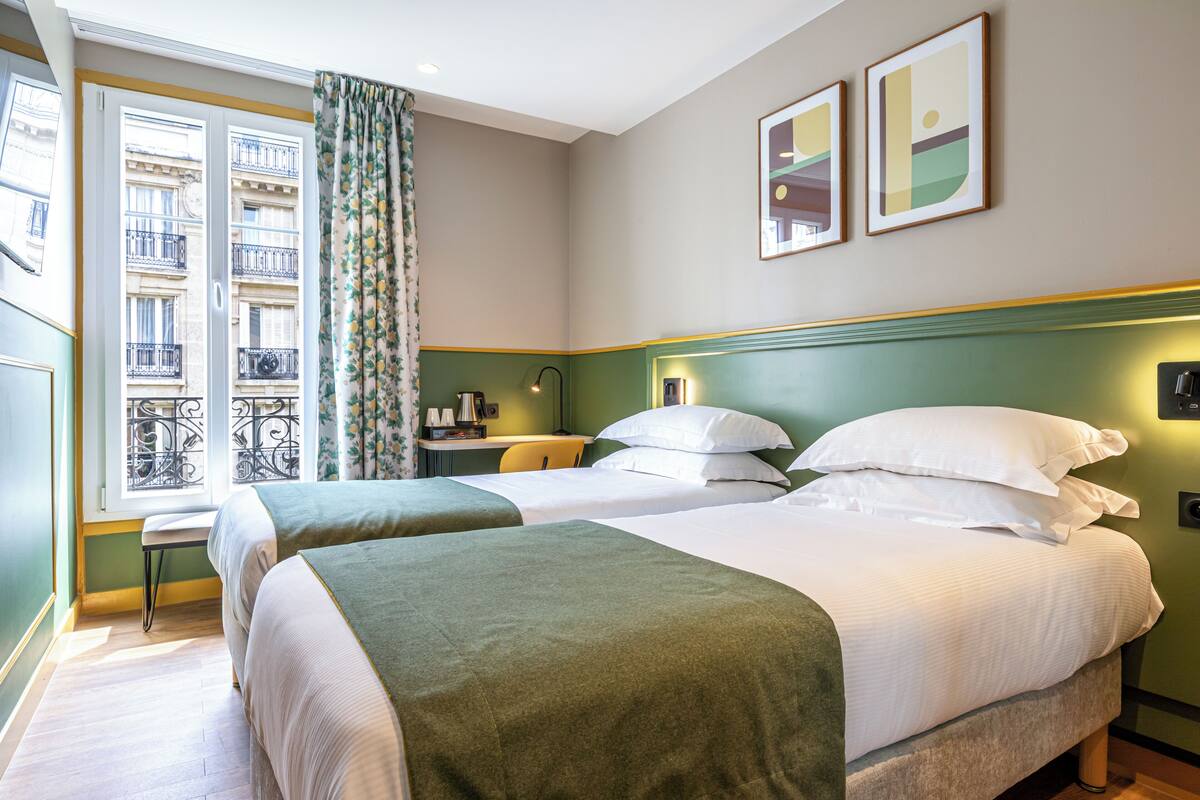 Quarto de hotel com duas camas, uma janela com vista para edifícios, uma escrivaninha e uma cadeira.