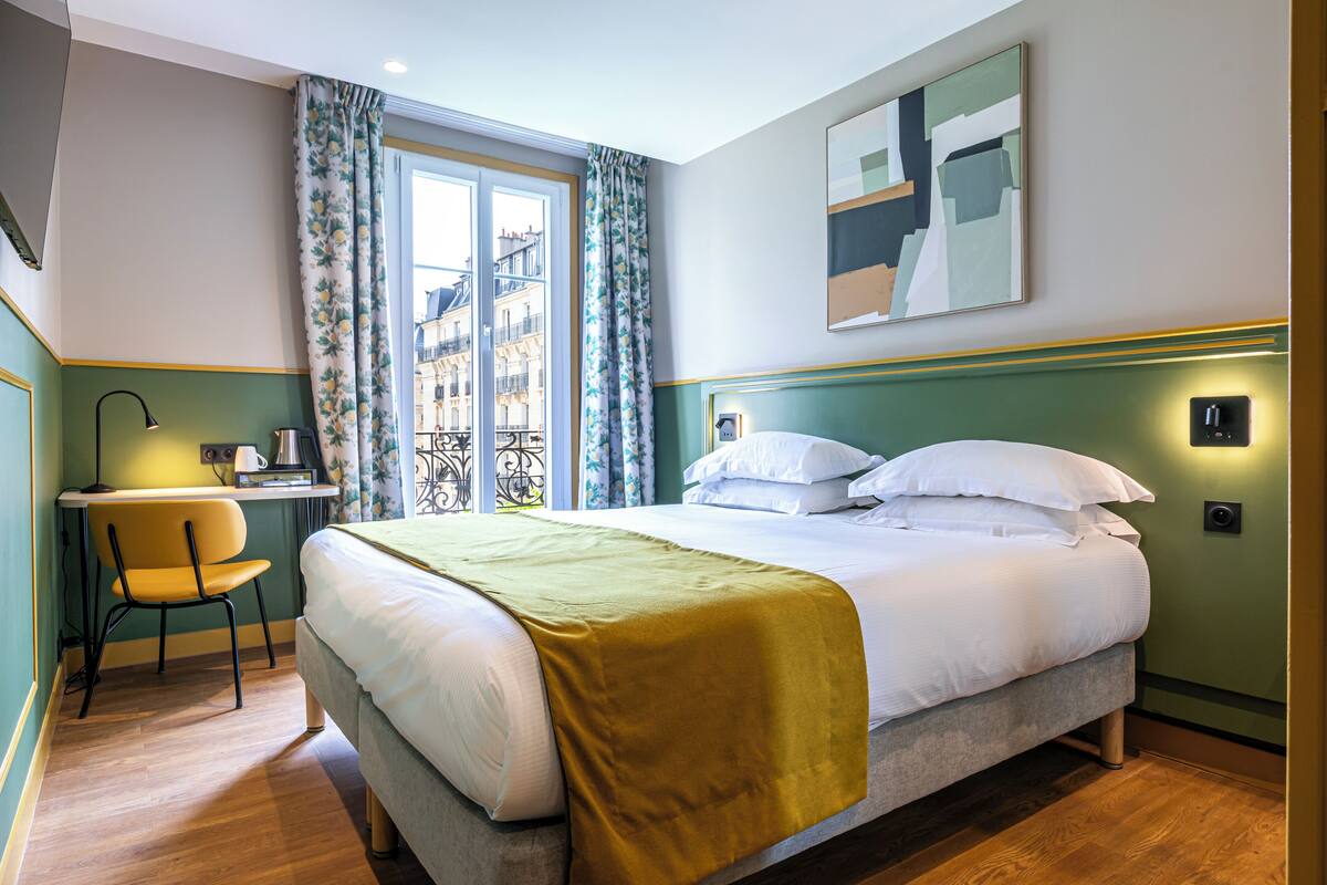 Quarto de hotel com cama, escrivaninha, cadeira, janela com cortinas e um quadro na parede.