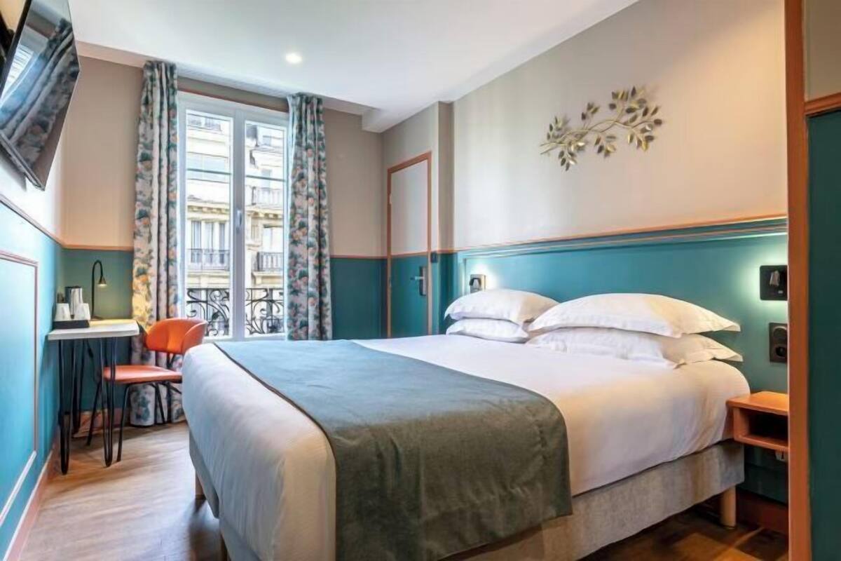 Quarto de hotel com cama, escrivaninha, cadeira e janela com cortinas.