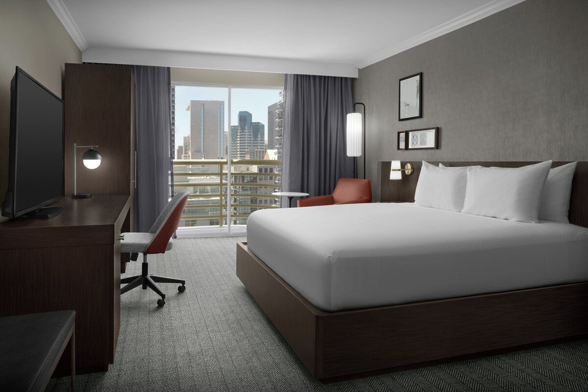 Quarto de hotel com uma cama grande, uma escrivaninha com cadeira, televisão, luminária e vista para a paisagem urbana.