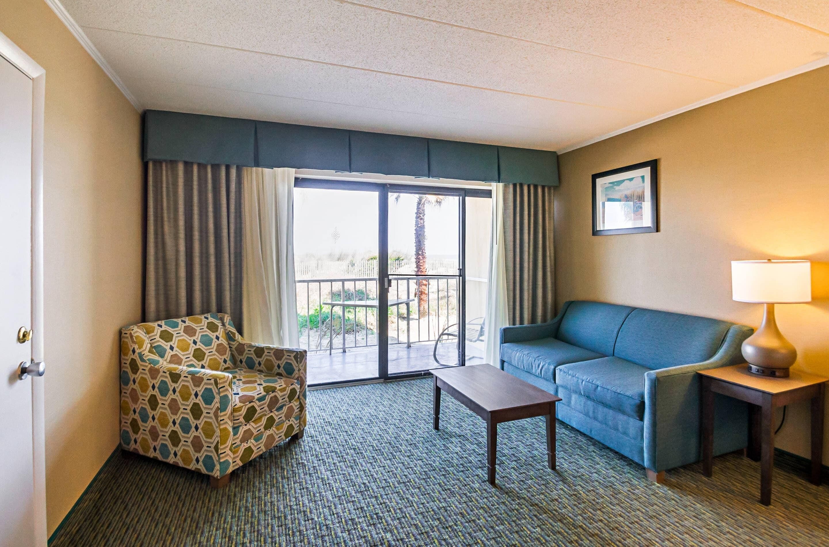 Quarto de hotel com uma poltrona estampada, um sofá azul, uma mesa pequena e uma luminária. Há uma porta de vidro deslizante que dá para uma varanda com vista para o exterior.