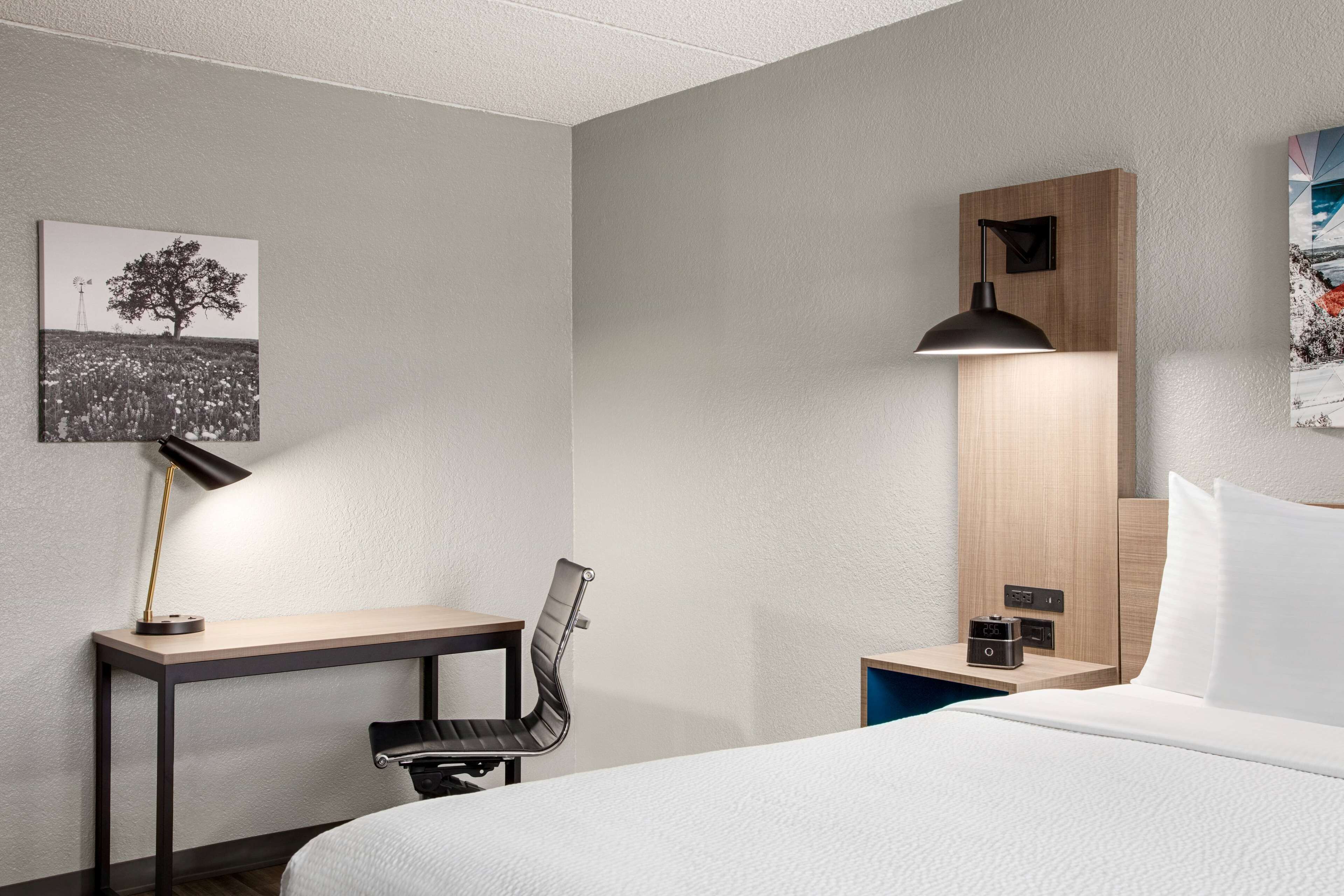 Quarto de hotel com cama, escrivaninha com cadeira, luminária, mesa de cabeceira com câmera e prateleira na parede.