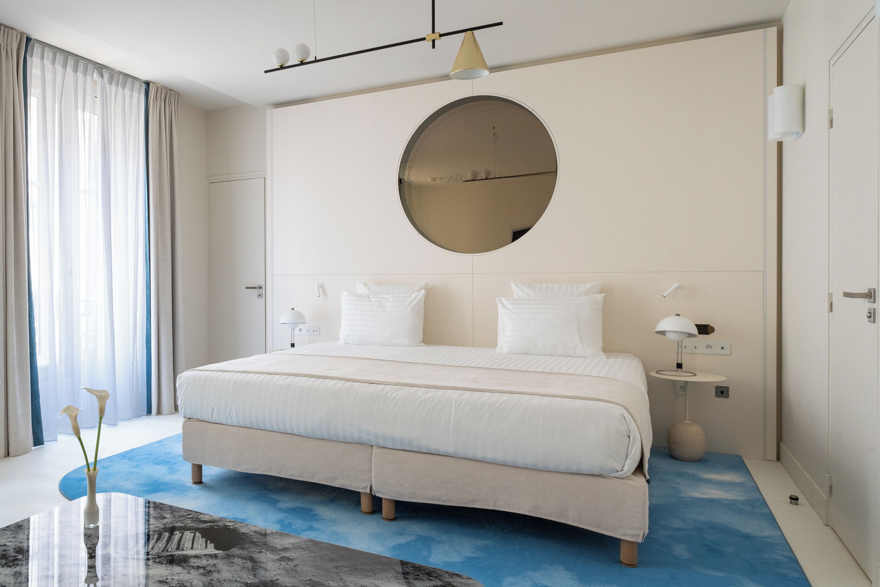 Quarto de hotel moderno com uma cama grande, um espelho redondo na parede, mesa de cabeceira com abajur e vista para o exterior através de cortinas leves.
