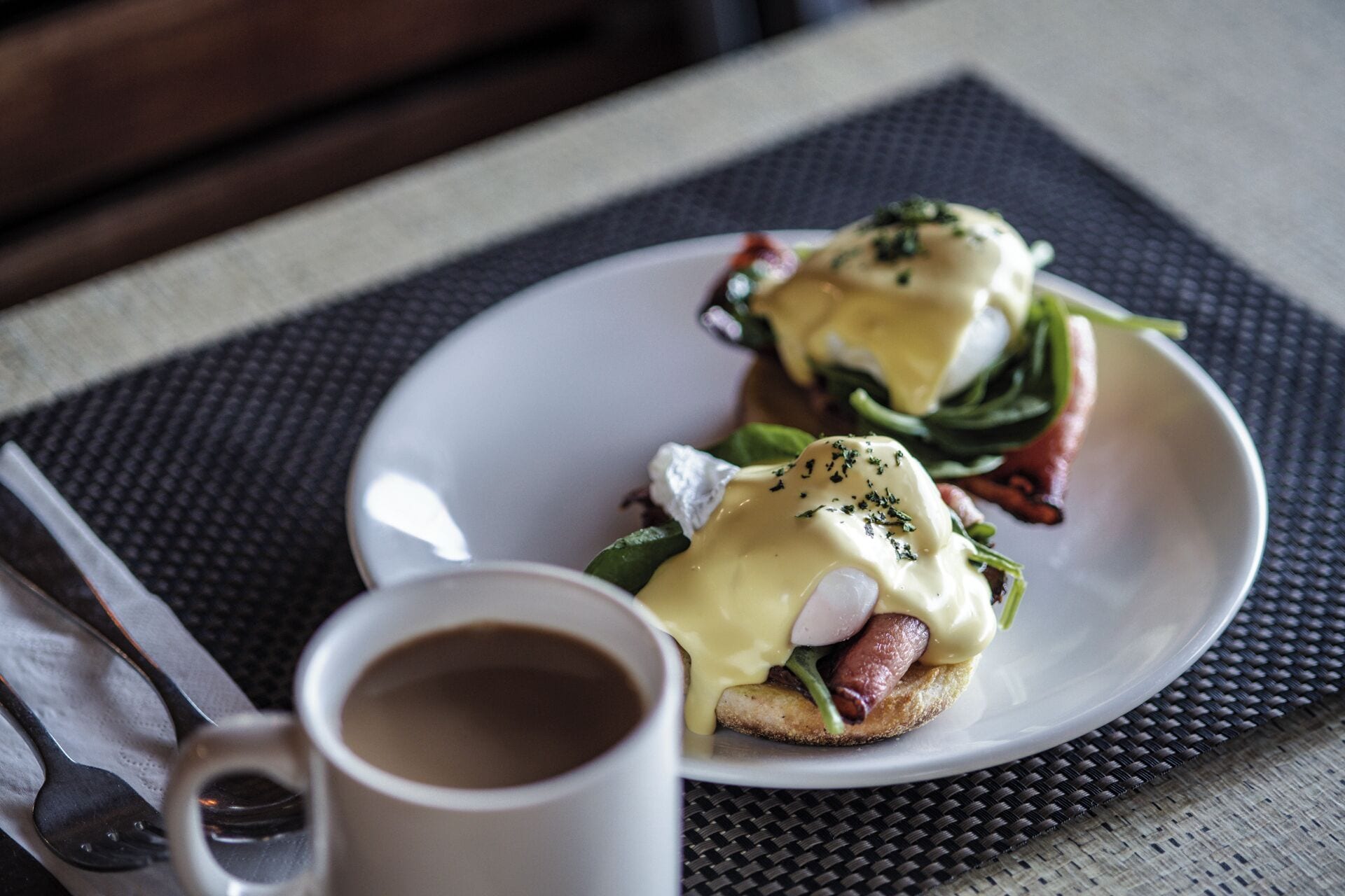 Um prato de ovos benedict com molho holandês, acompanhado de uma xícara de café sobre uma mesa com sousplat.