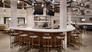 Uma área de bar moderna com balcão curvo, vários bancos e uma variedade de garrafas e copos em prateleiras.