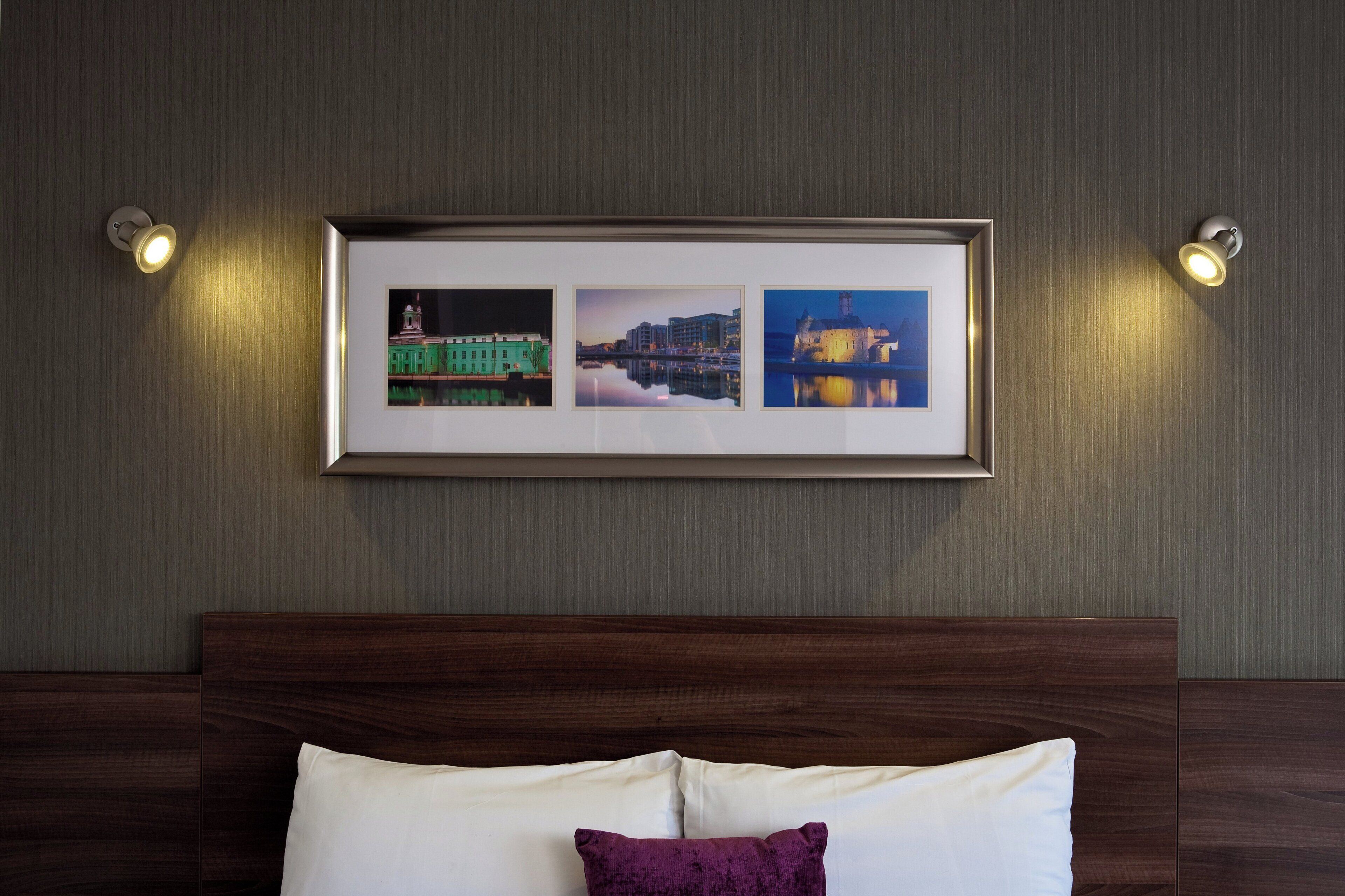 Quadro com três paisagens urbanas numa parede acima de uma cama com cabeceira de madeira e almofadas brancas.